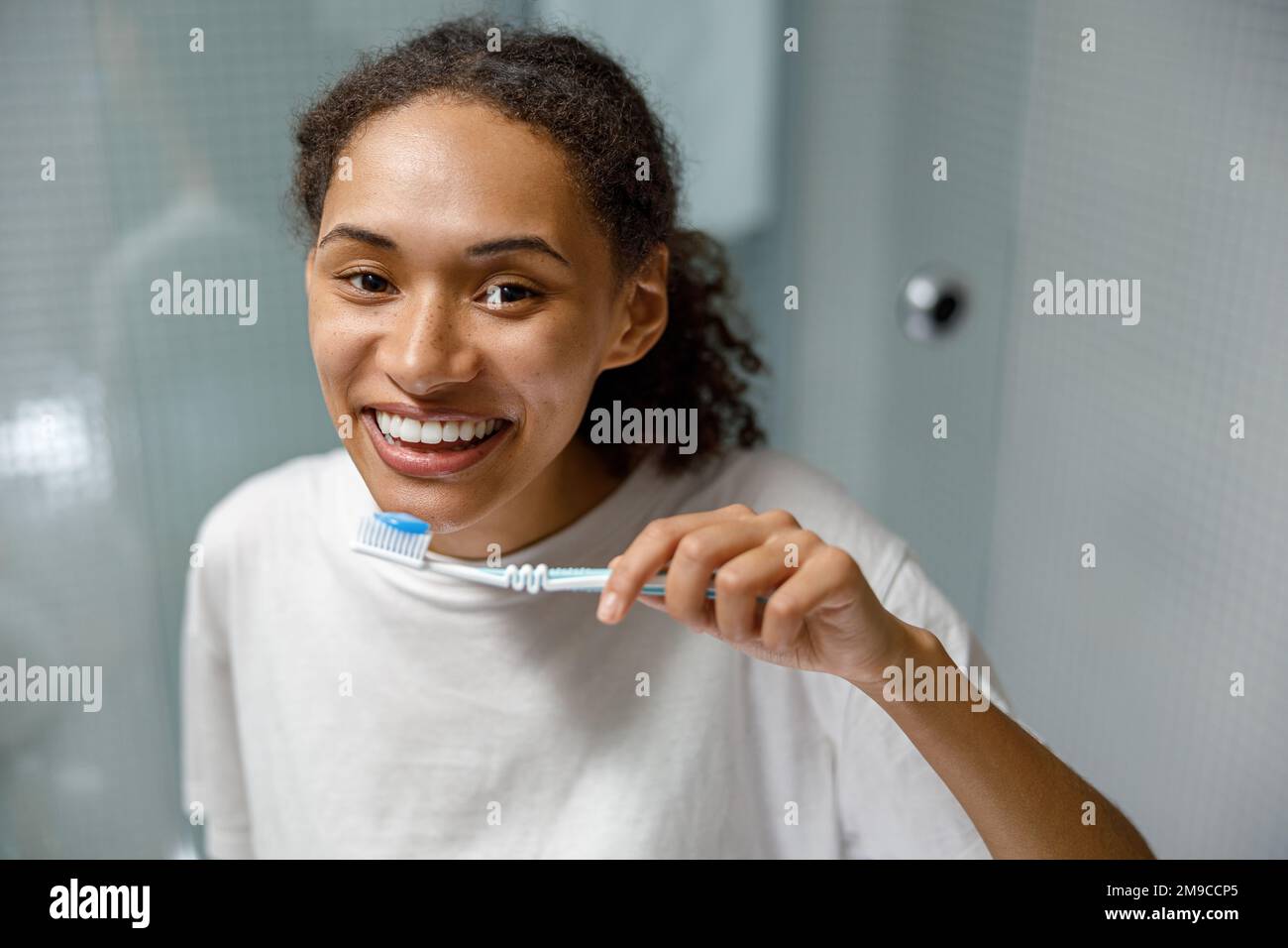 Smiling woman brushing teeth in bathroom and looking at camera. Morning ...