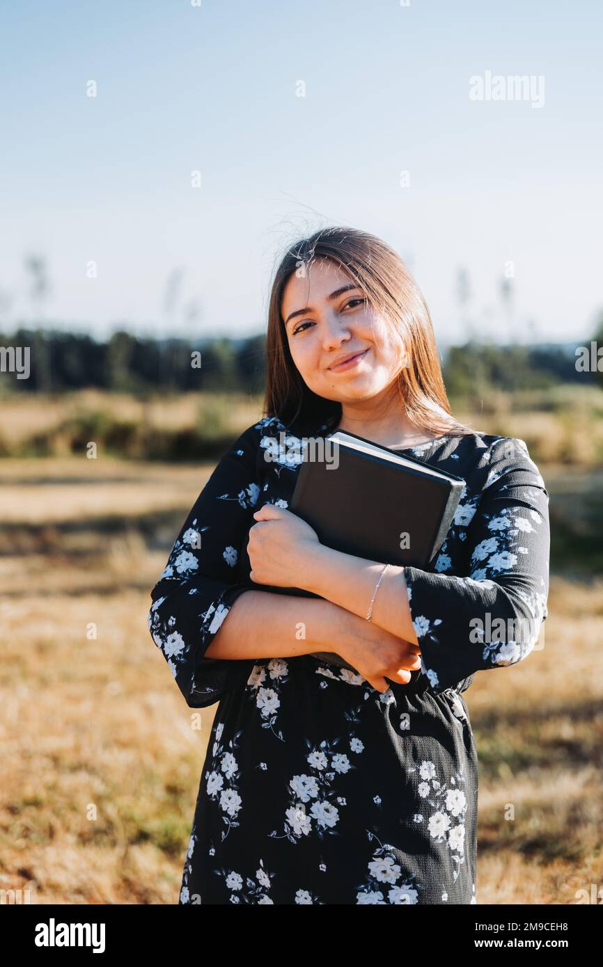 Young smiling religious girl holding a bible under her arm in the field ...