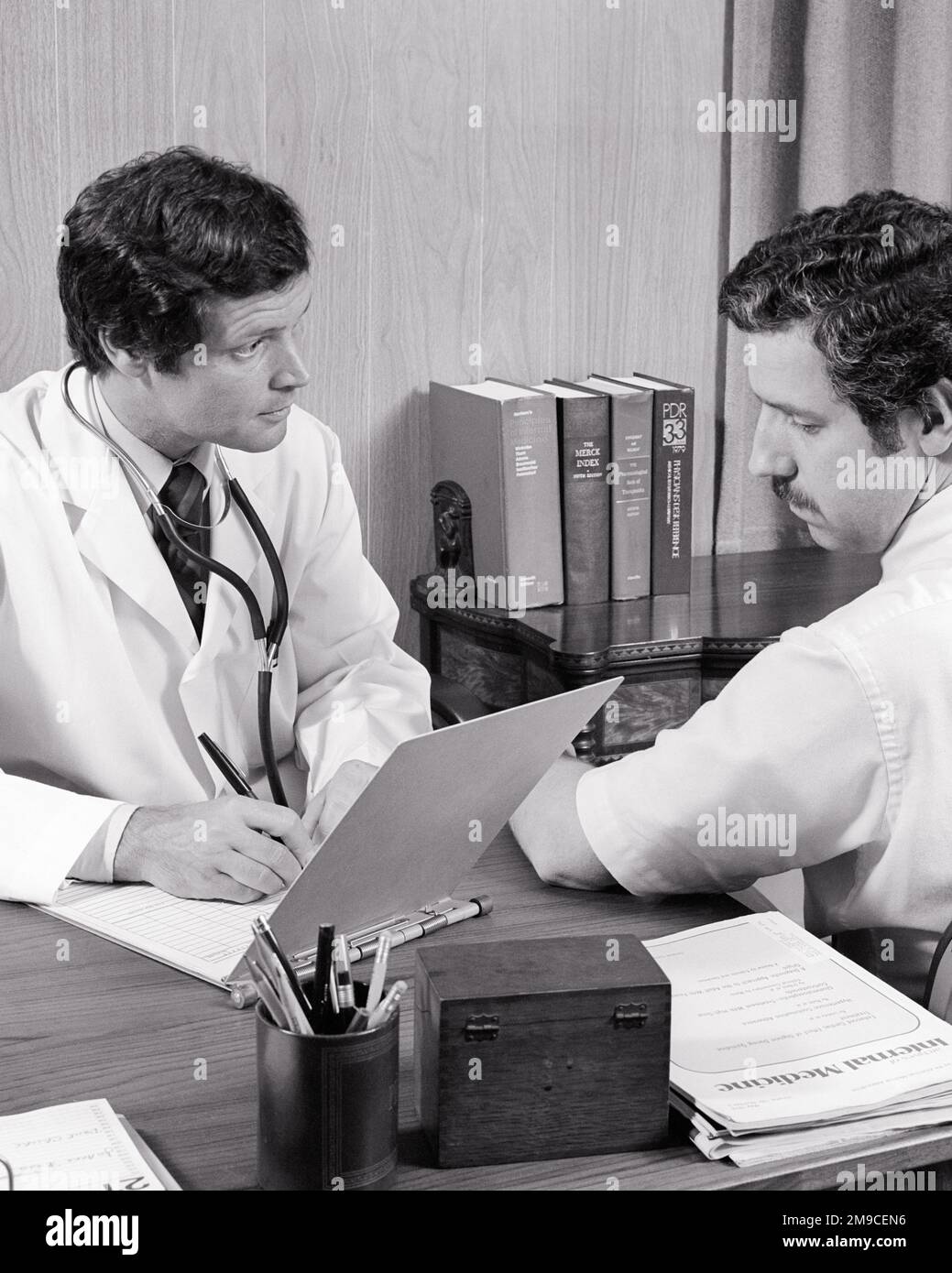 1970s DOCTOR WRITING IN A CHART WHILE CONSULTING WITH A MALE PATIENT IN THE DOCTOR’S OFFICE ...