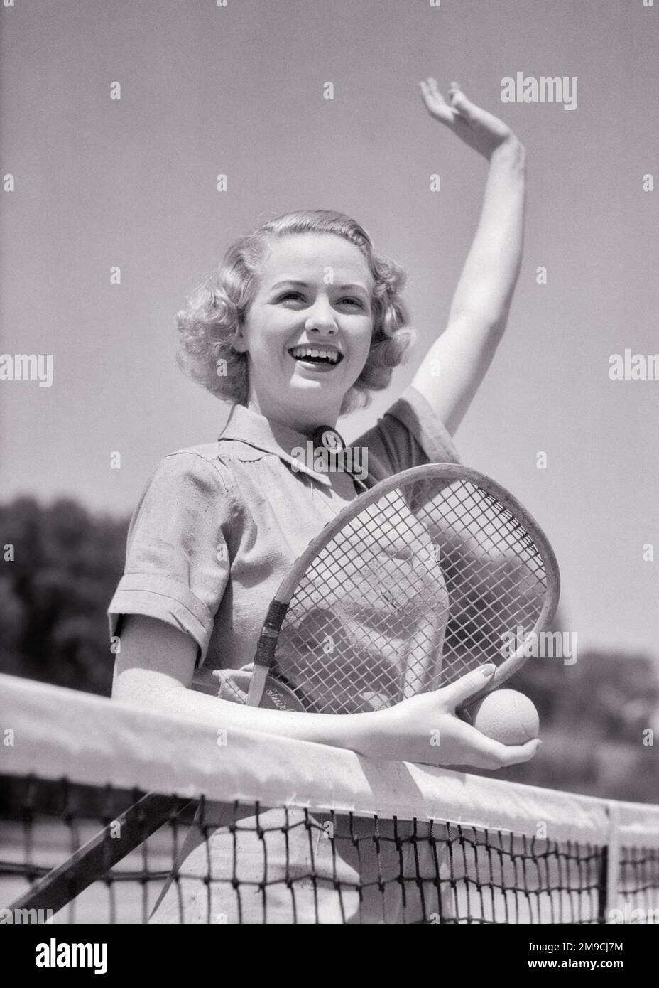 1930s BLONDE WOMAN SMILING WAVING HAND STANDING BY TENNIS NET HOLDING ...