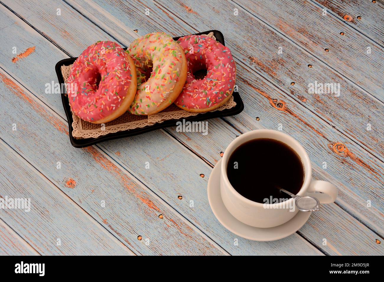 Three donuts on a plate and a cup of hot black coffee on a wooden table ...