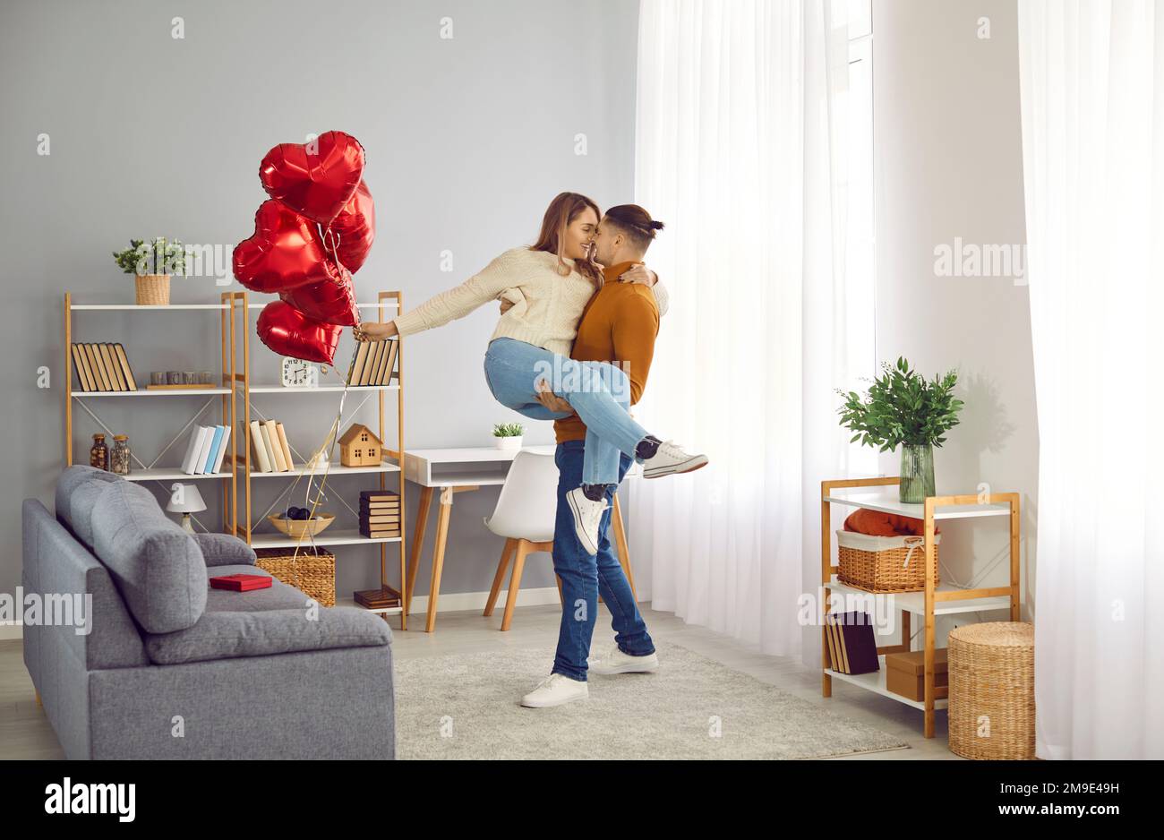 Boy holding a girl in his arms indoor. Young happy couple in love with red heart shaped ballons ...