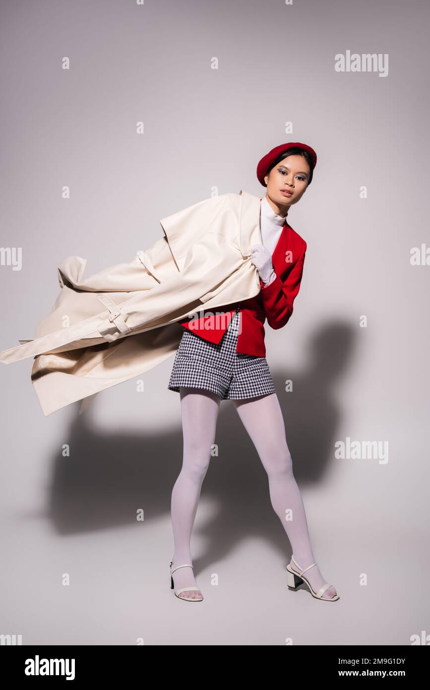 Trendy asian woman in beret wearing trench coat on grey background ...