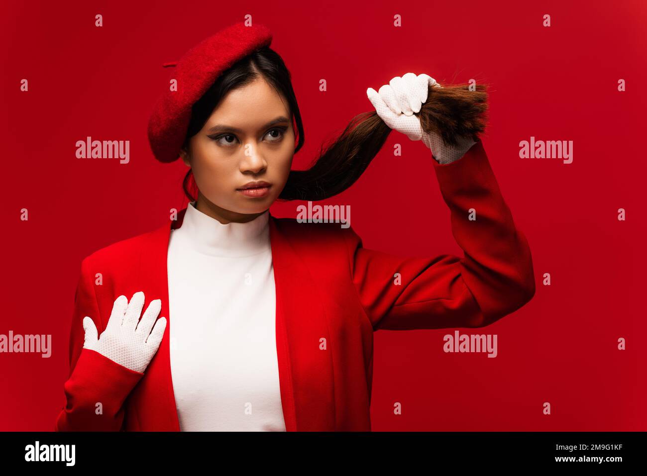 Pretty asian woman in beret and jacket touching hair isolated on red ...