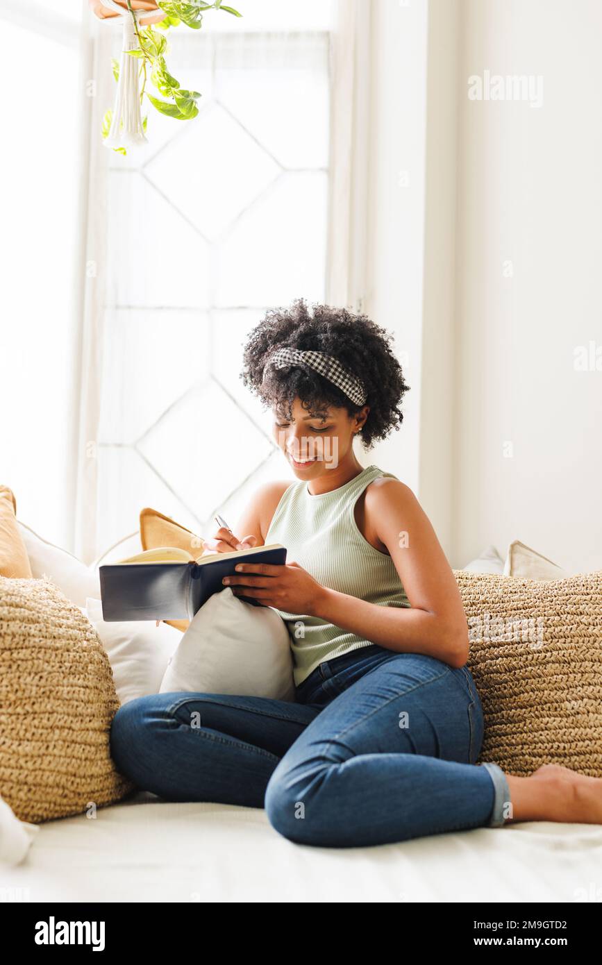 Smiling biracial young woman with afro hair writing in diary while ...