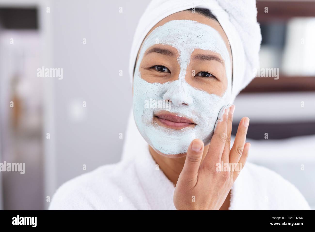 Portrait of happy asian woman applying beauty face mask in bathroom ...