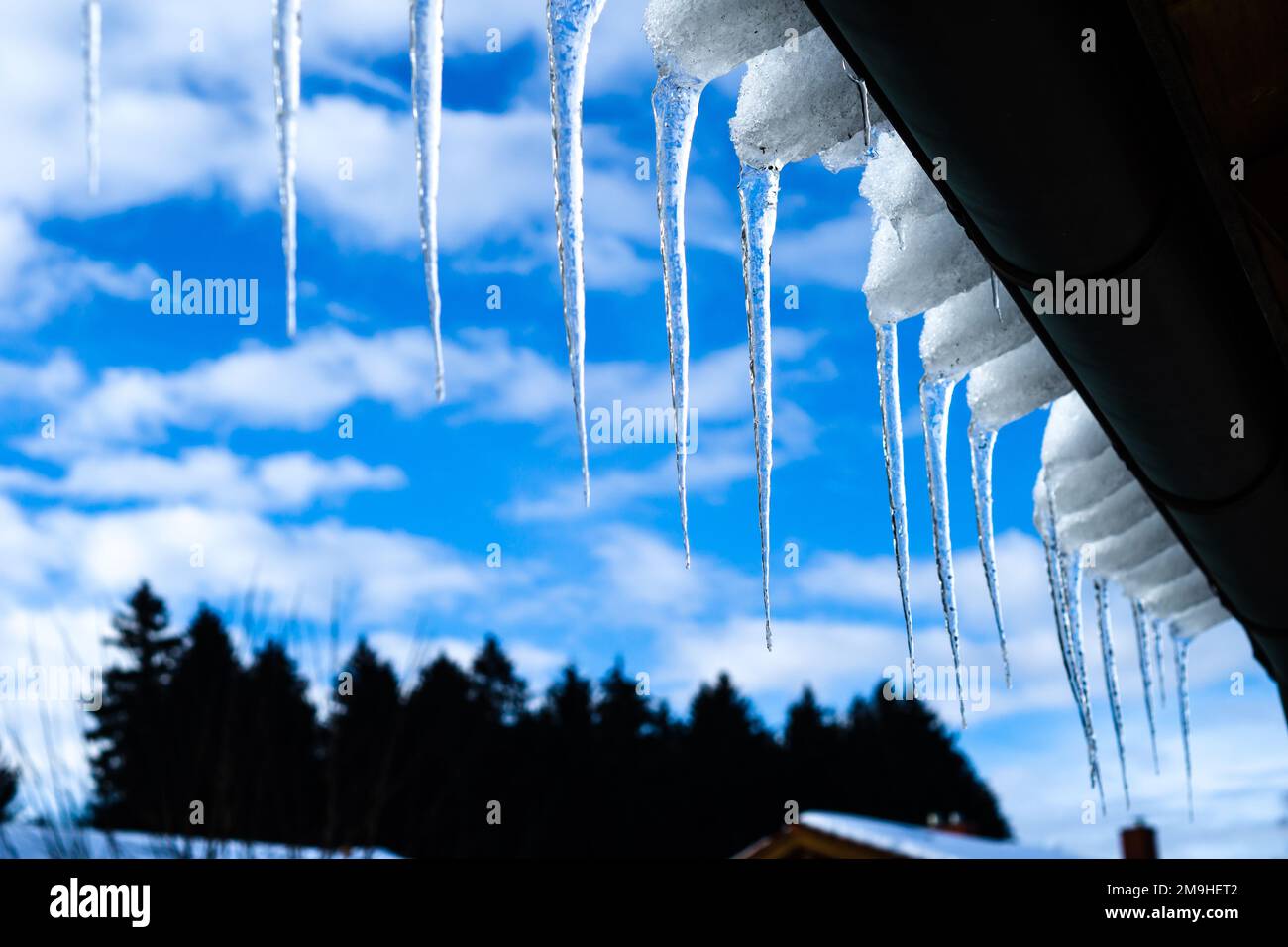 A closeup of ice dams hanging from a roof with cloudy sky blurred ...