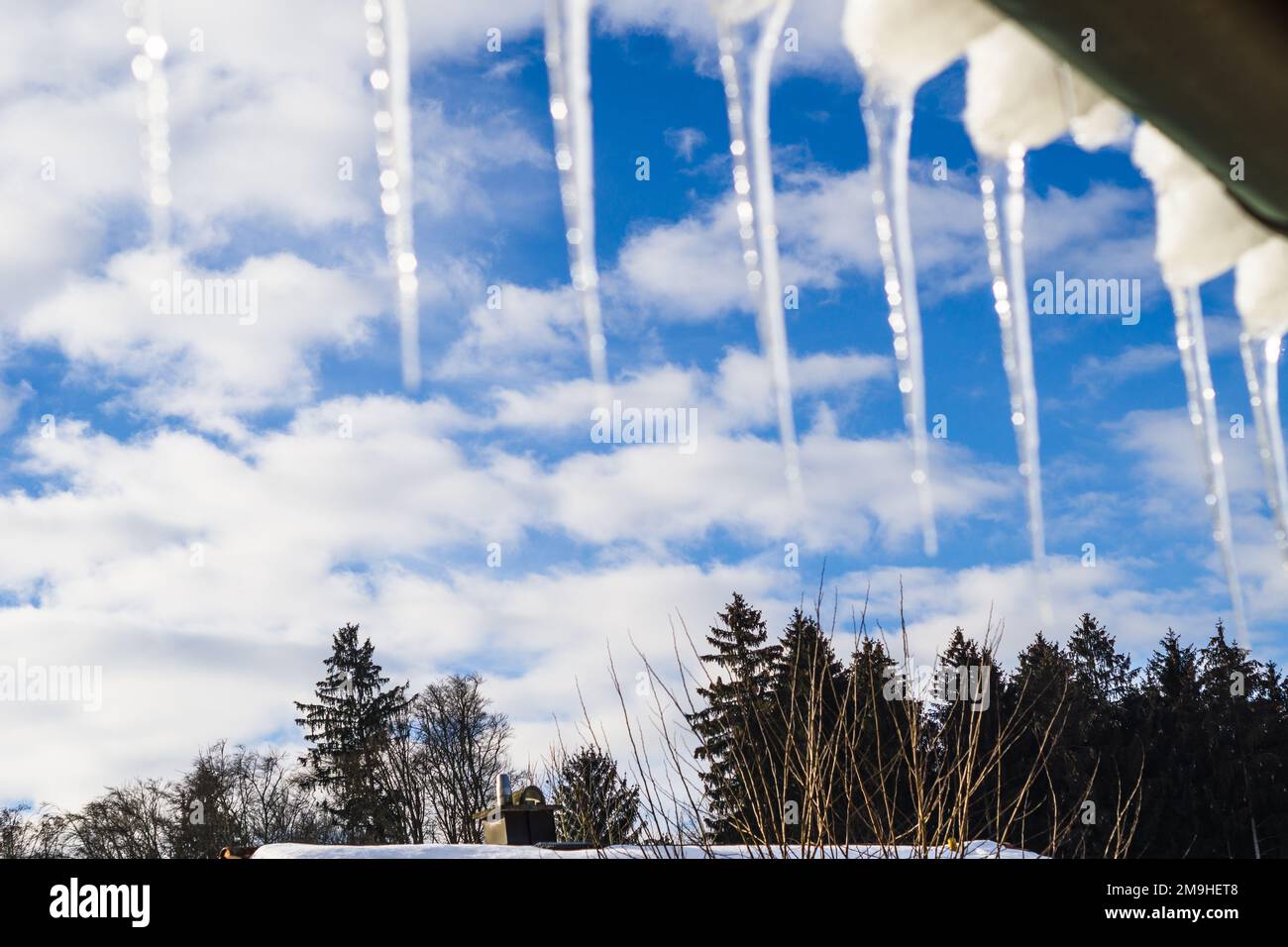 A closeup of ice dams hanging from a roof with cloudy sky blurred ...