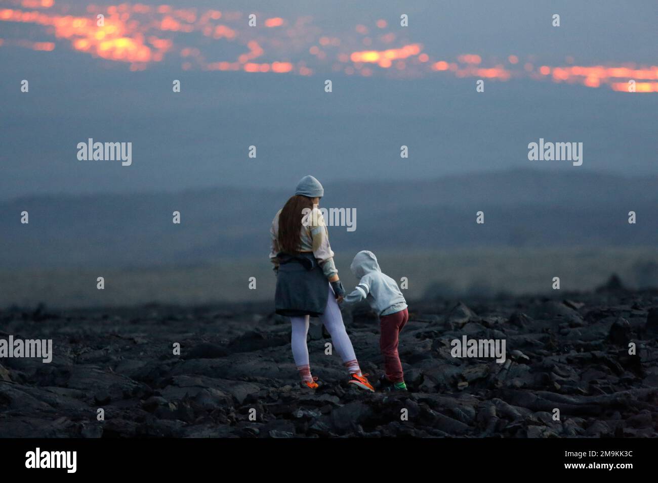 A woman and child walk through an old lava field as fresh lava erupts ...