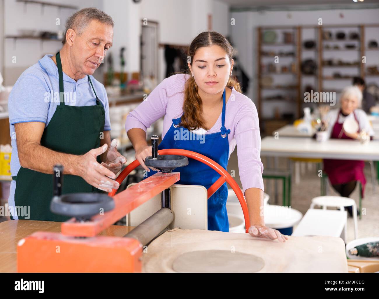 Elderly potter teaches woman how to roll clay on a craft machine clay ...