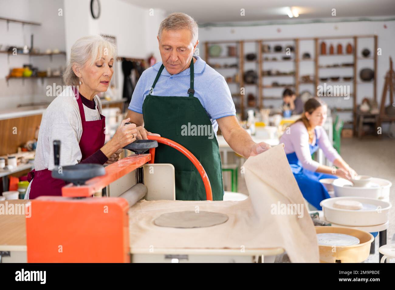 Elderly potter teaches woman how to roll clay on a craft machine clay ...