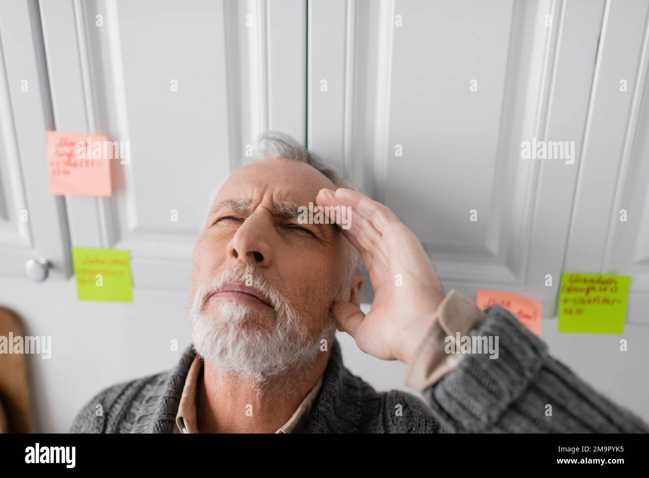 senior man with closed eyes and hand near head standing near blurred ...