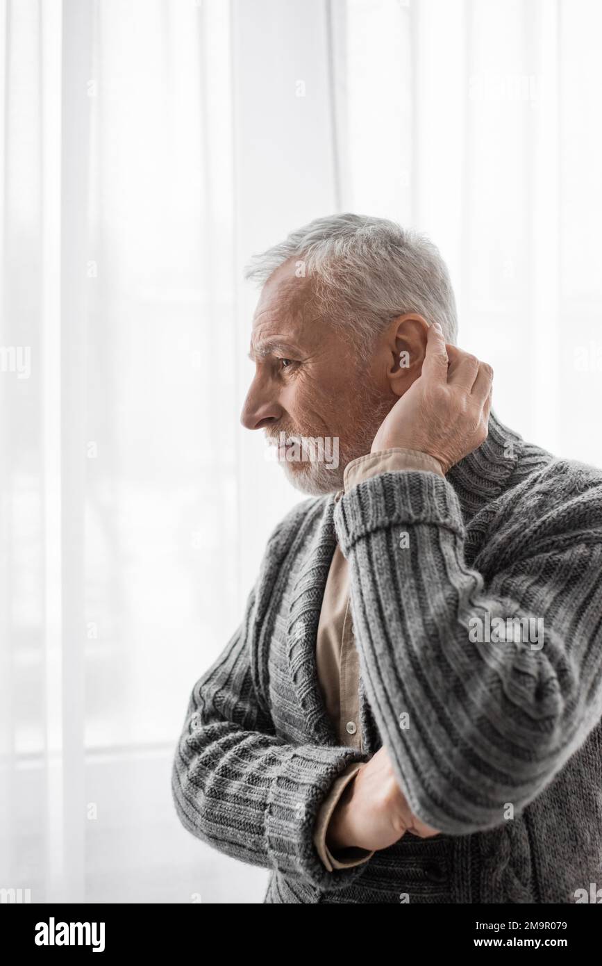 senior and thoughtful man in knitted cardigan looking away near window ...