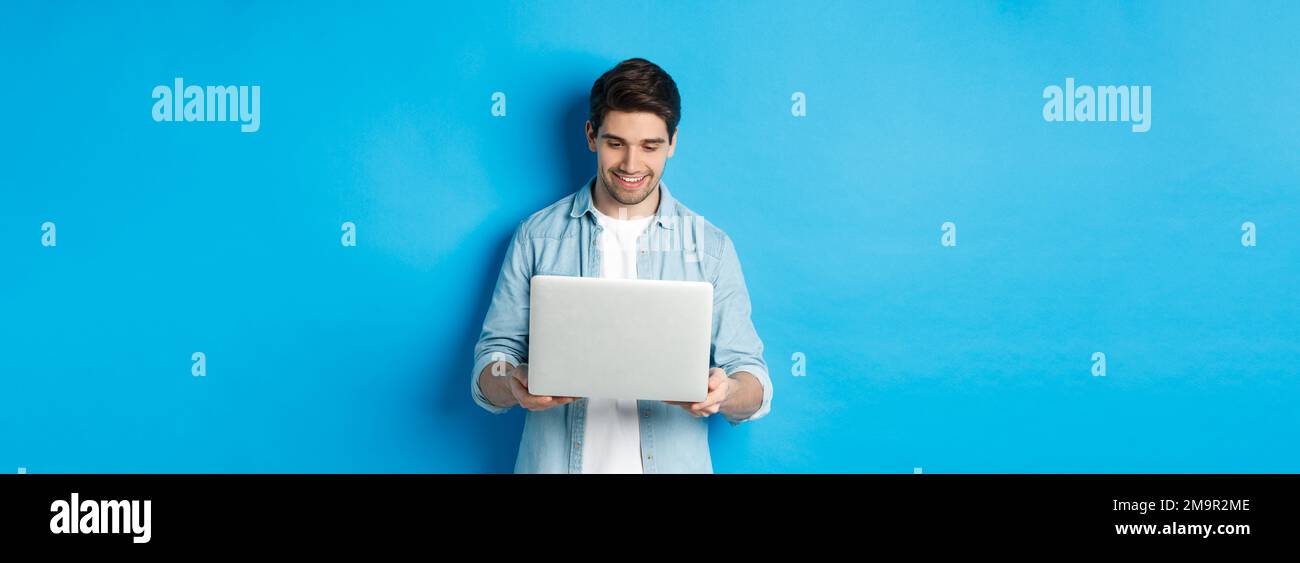 Handsome man working on laptop, smiling and looking at screen satisfied ...