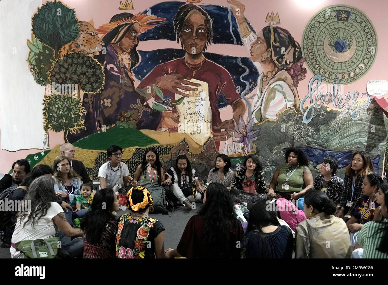 Indigenous people hold hands in a circle backdropped by a mural by ...