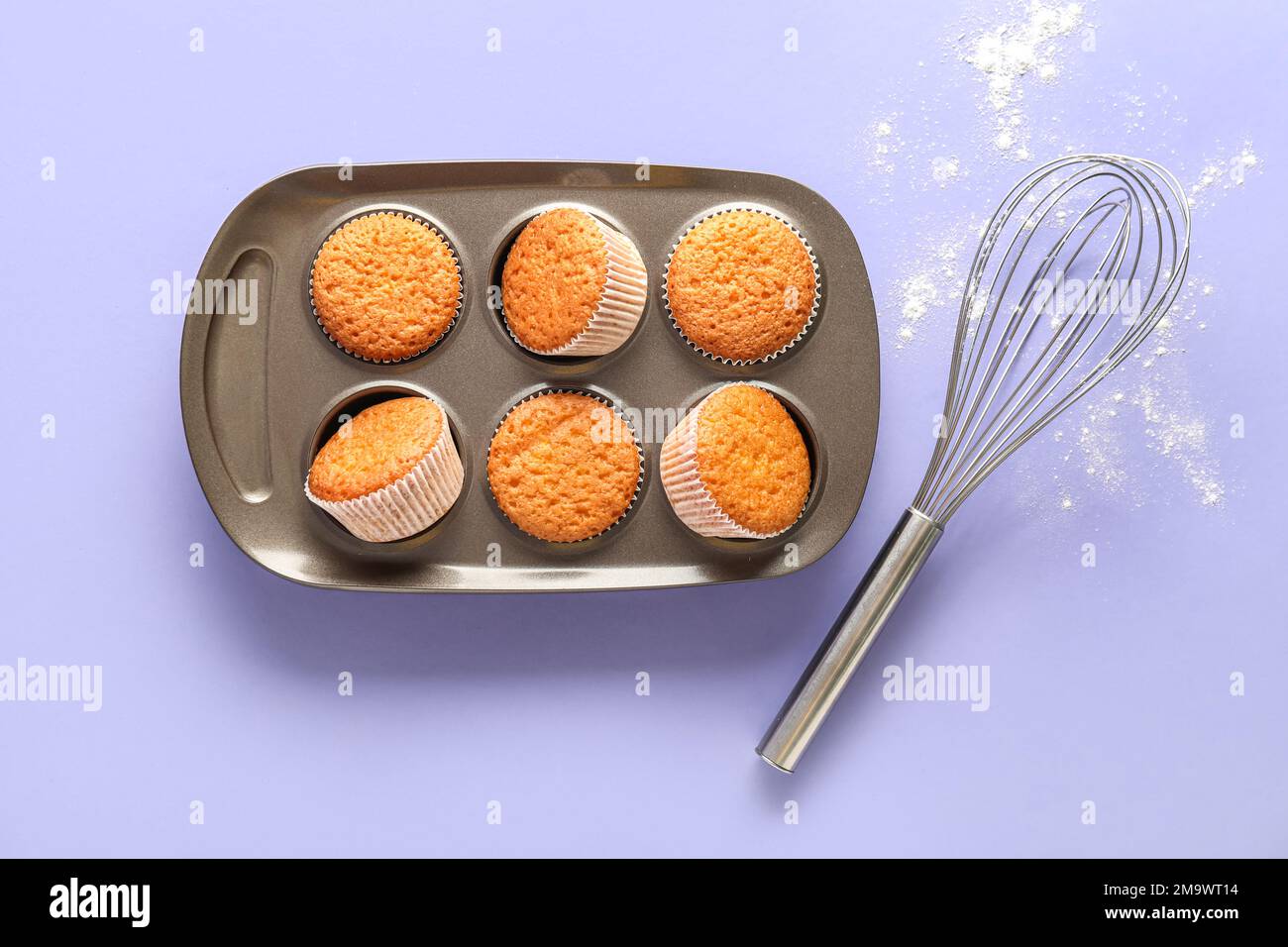Baking tray with muffins, whisk and scattered flour on lilac background ...