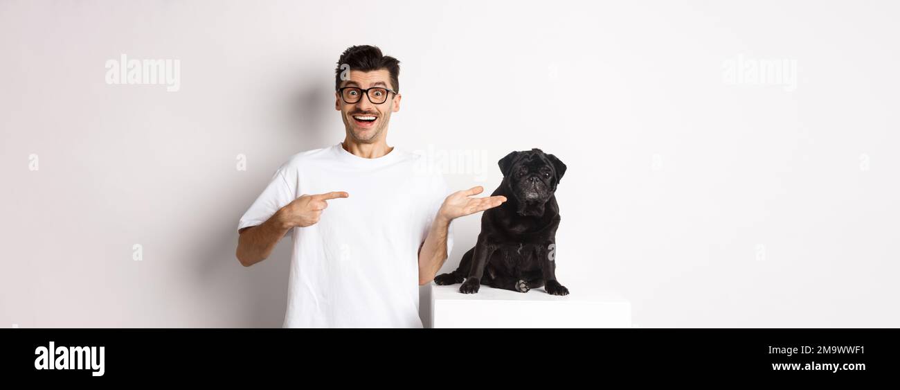 Cheerful young man pointing finger at his dog, showing small cute black ...