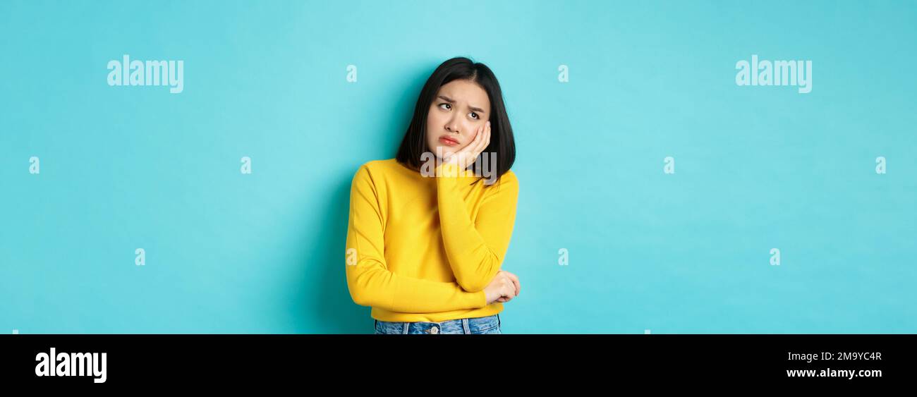 Sad and bored asian teenage girl leaning on palm, looking at upper left ...