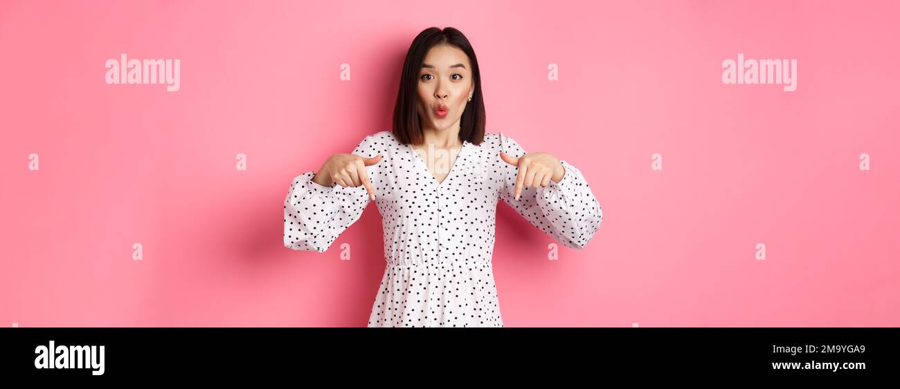 Impressed asian woman in dress pointing fingers down, look there ...