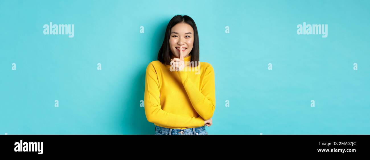 Cheerful asian girl asking to be queit, shushing at camera and smiling ...