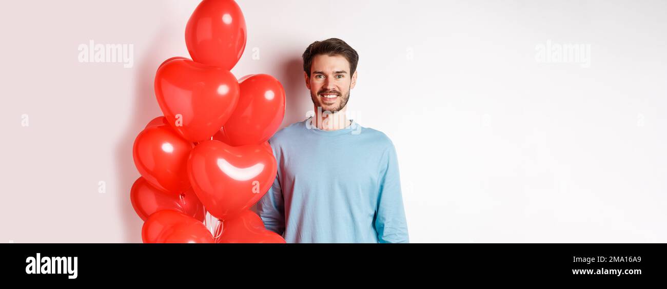 Smiling young man standing with heart balloons and looking happy ...