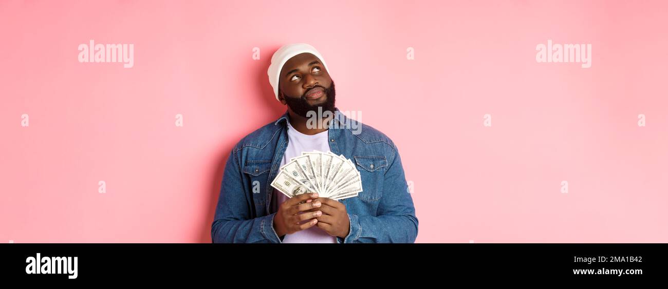 Dreamy african-american man thinking about shopping, holding money ...