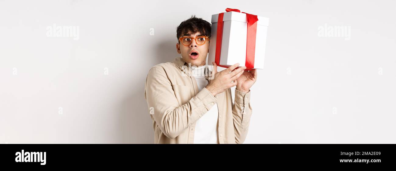 Holidays concept. Funny young man in glasses shaking gift box ...