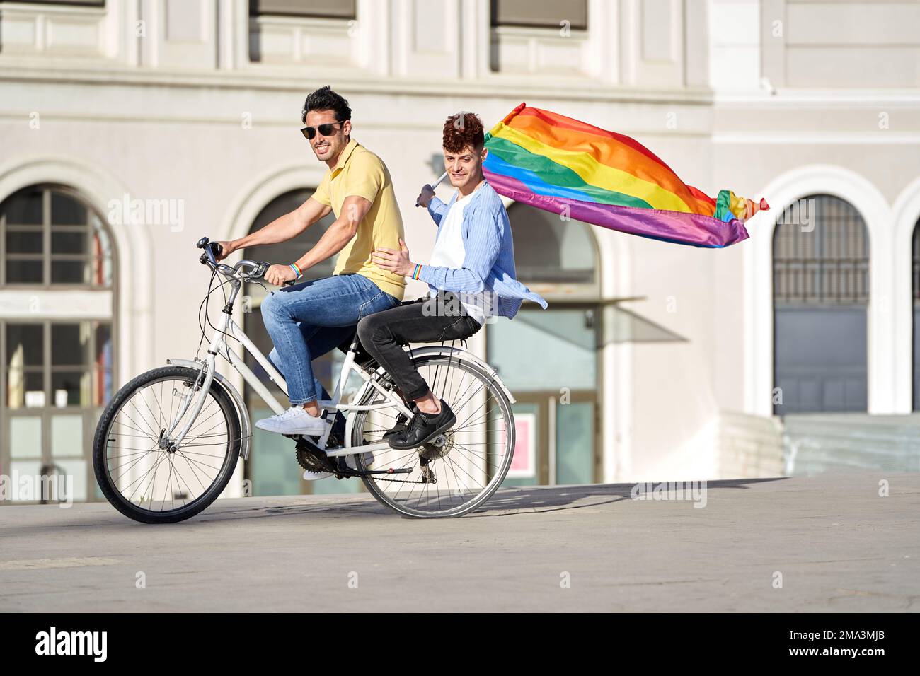 Side view of Young gay couple holding a gay pride flag while riding a ...