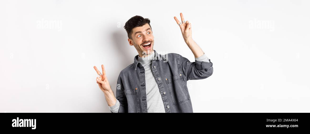 Funny young man showing v-signs and smiling at left side, looking at ...