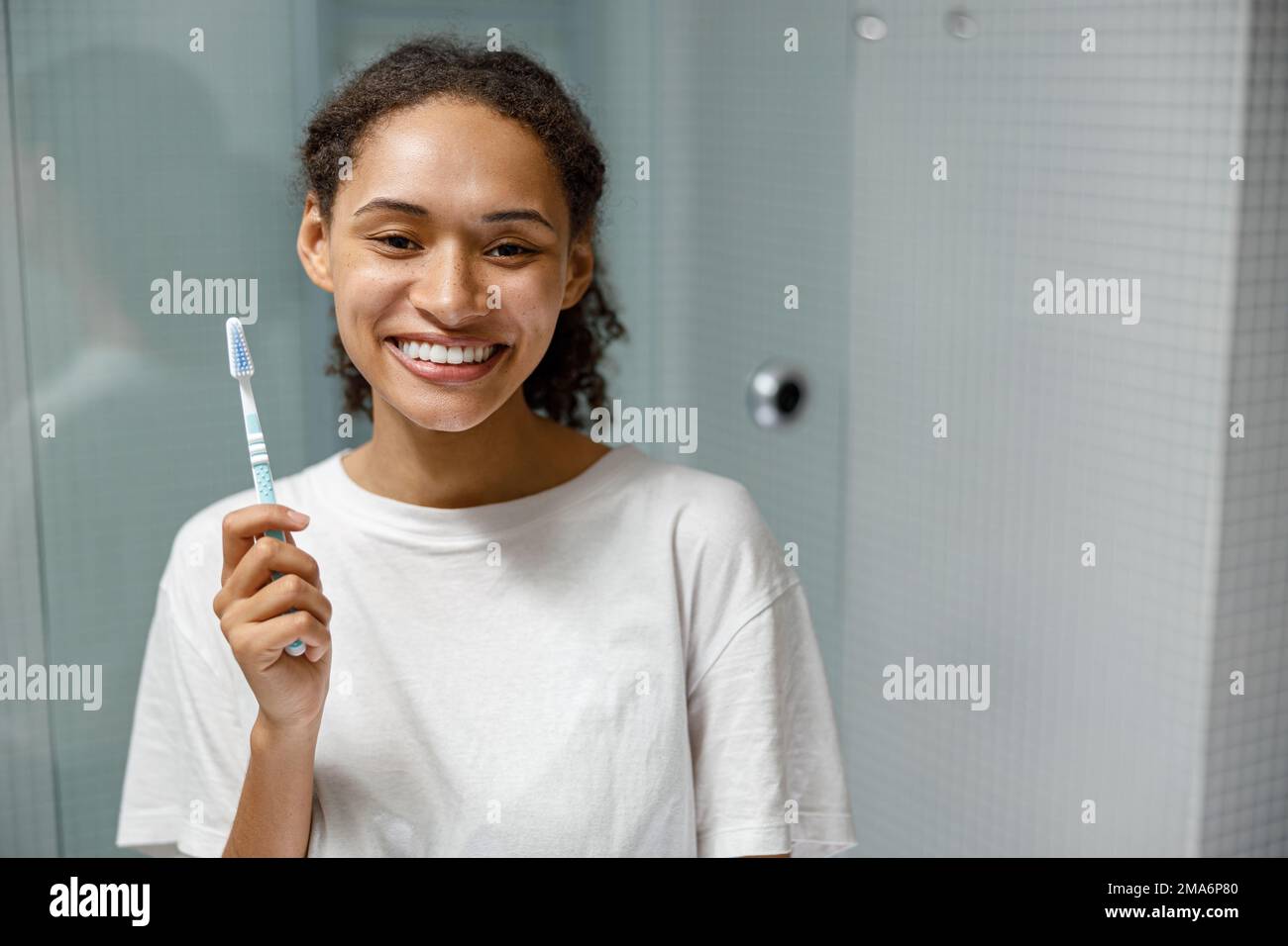 Smiling woman brushing teeth in bathroom and looking at camera. Morning ...