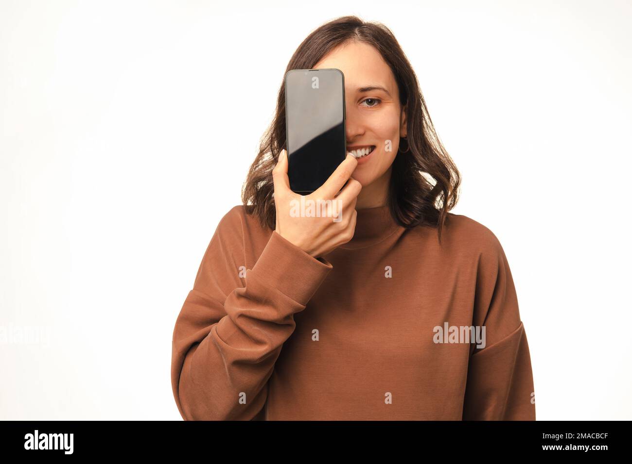 Smiling woman is hiding half of her face behind phone while showing its ...