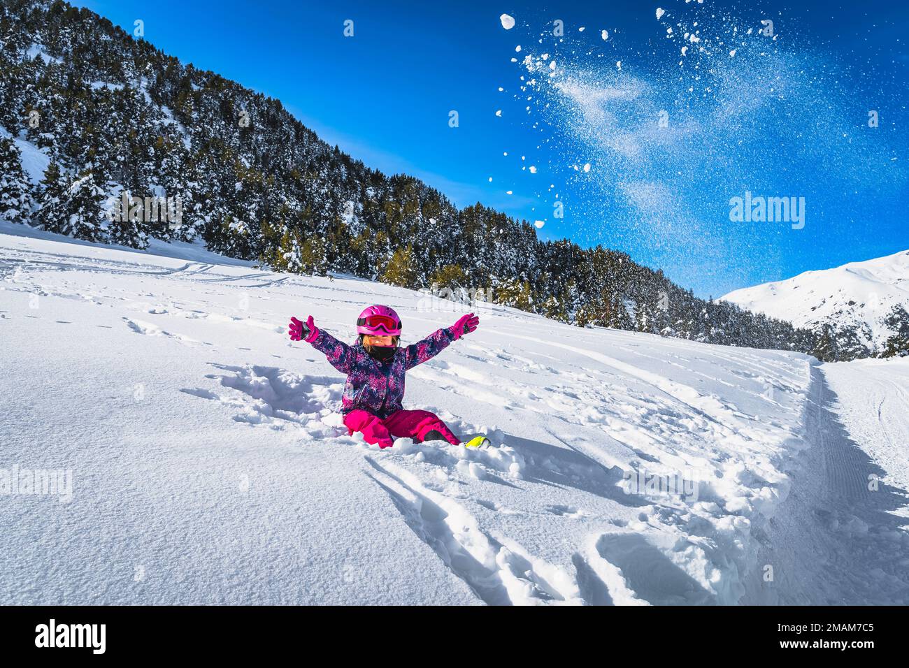 Smiling girl sitting on snow and throwing snow powder up to the air ...