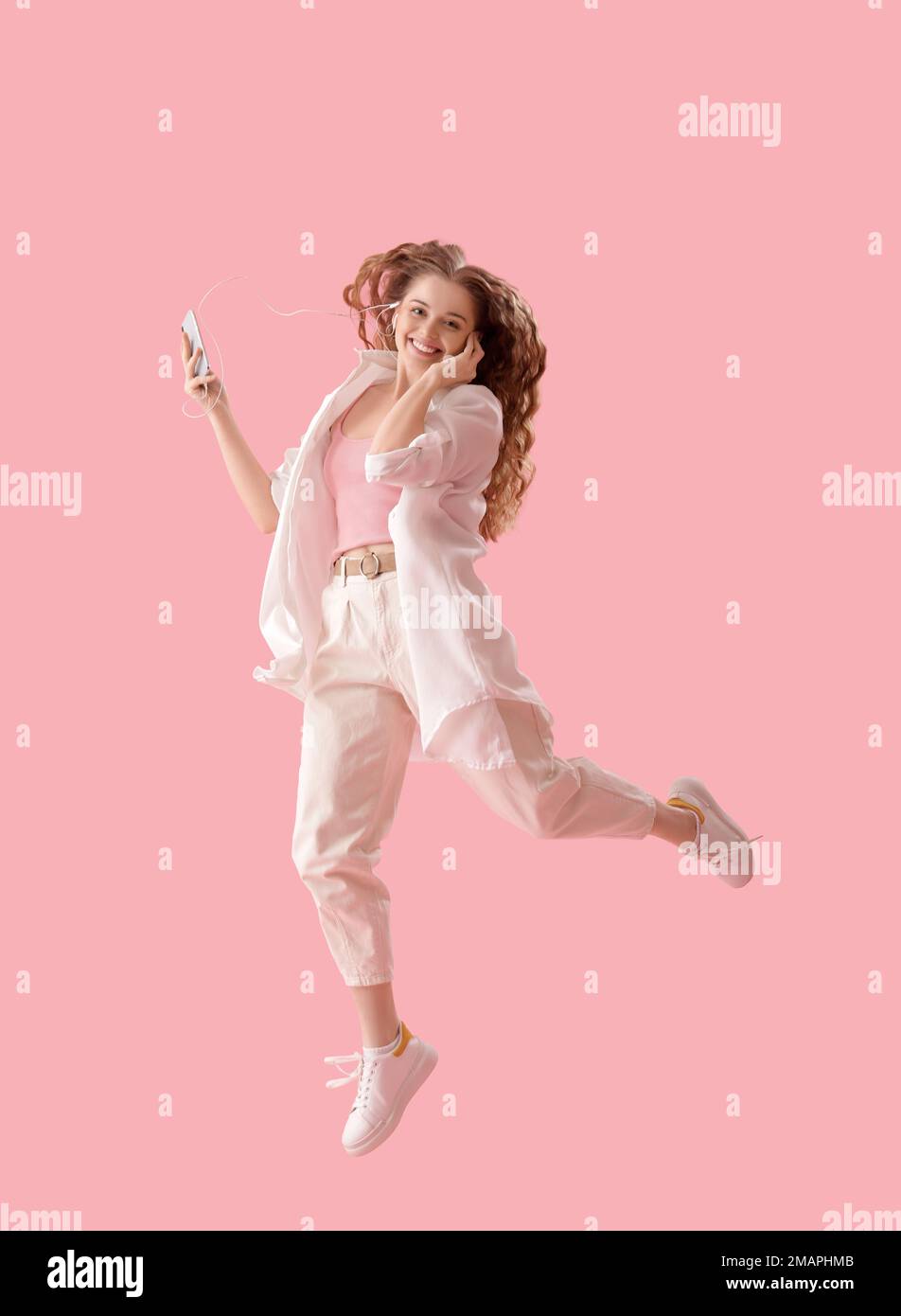 Young woman in earphones with mobile phone dancing on pink background ...
