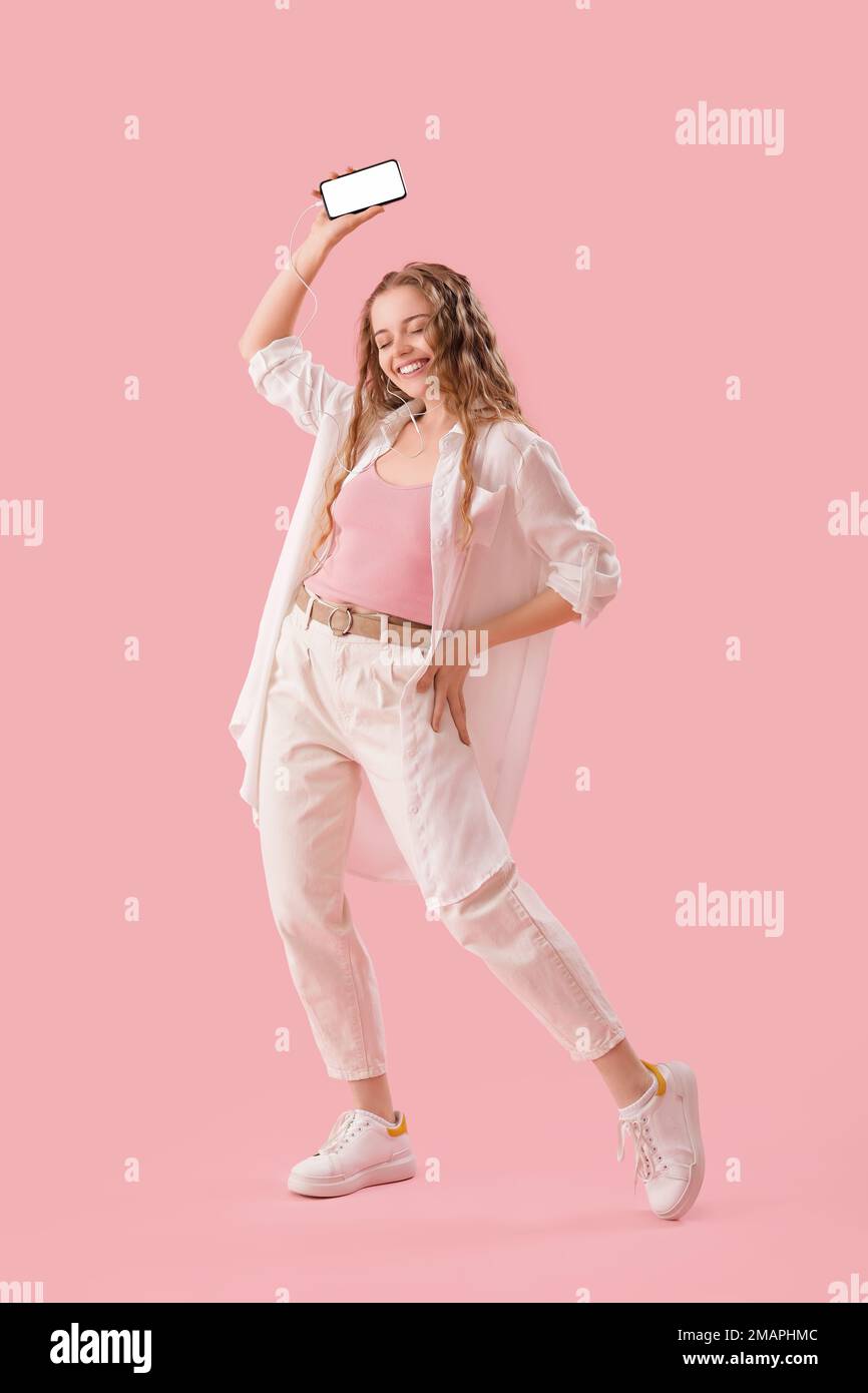 Young woman in earphones with mobile phone dancing on pink background ...