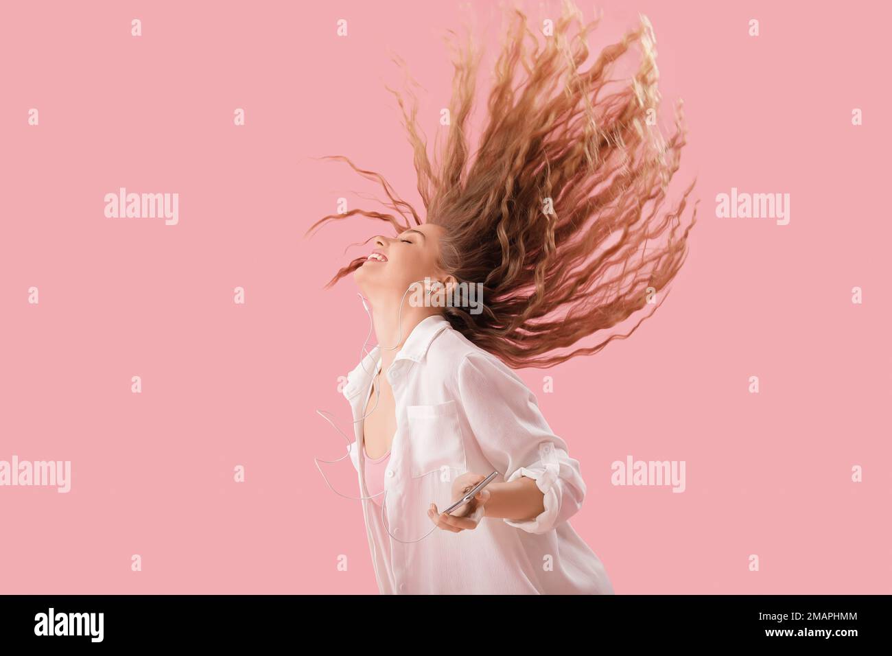 Young woman in earphones with mobile phone dancing on pink background ...