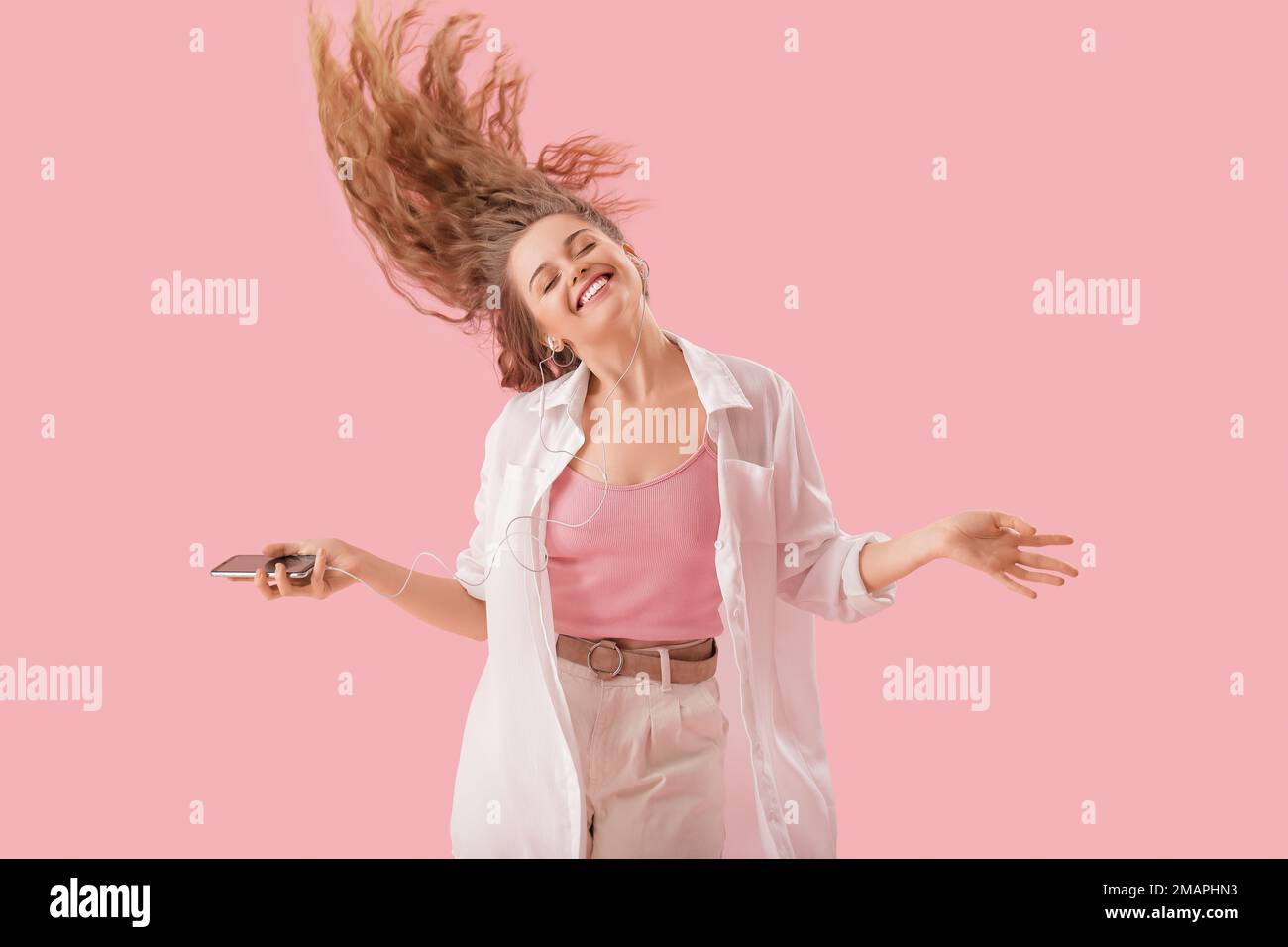 Young woman in earphones with mobile phone dancing on pink background ...