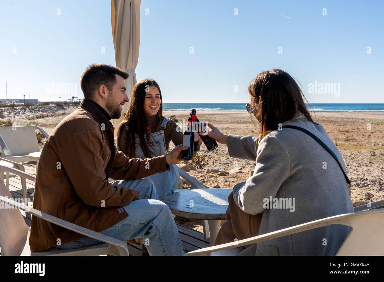 Young friends in warm clothes sitting at table and clinking bottles of ...
