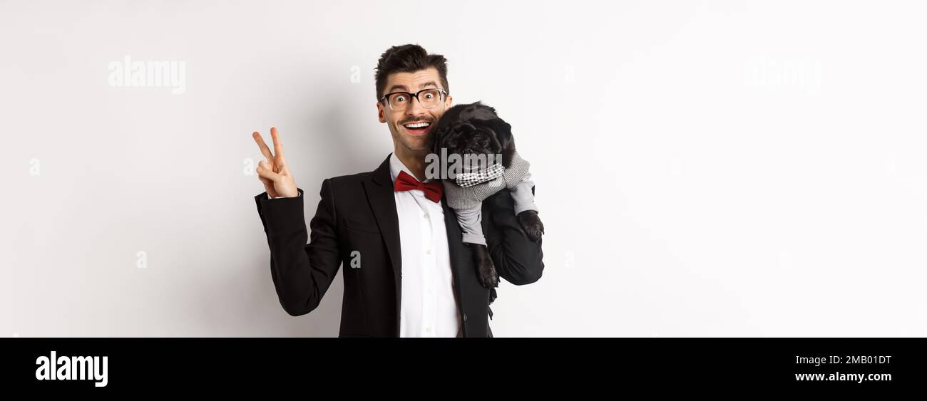 Cheerful young man in suit and glasses taking photo with cute black pug ...