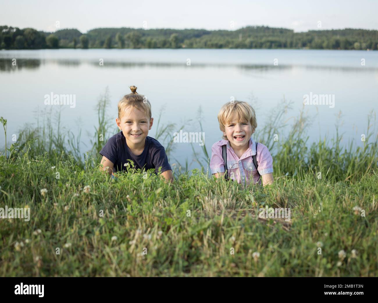 Two happy children lie on their stomachs in the grass near the lake ...