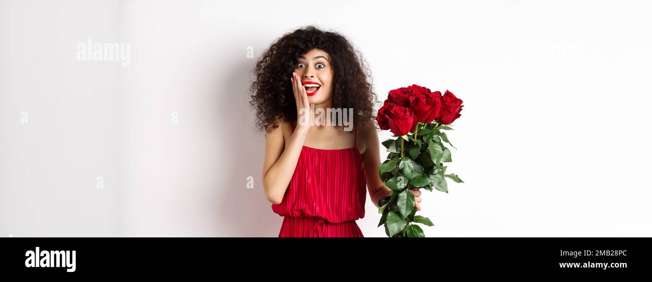 Excited curly woman in red dress, receive bouquet of roses and look ...