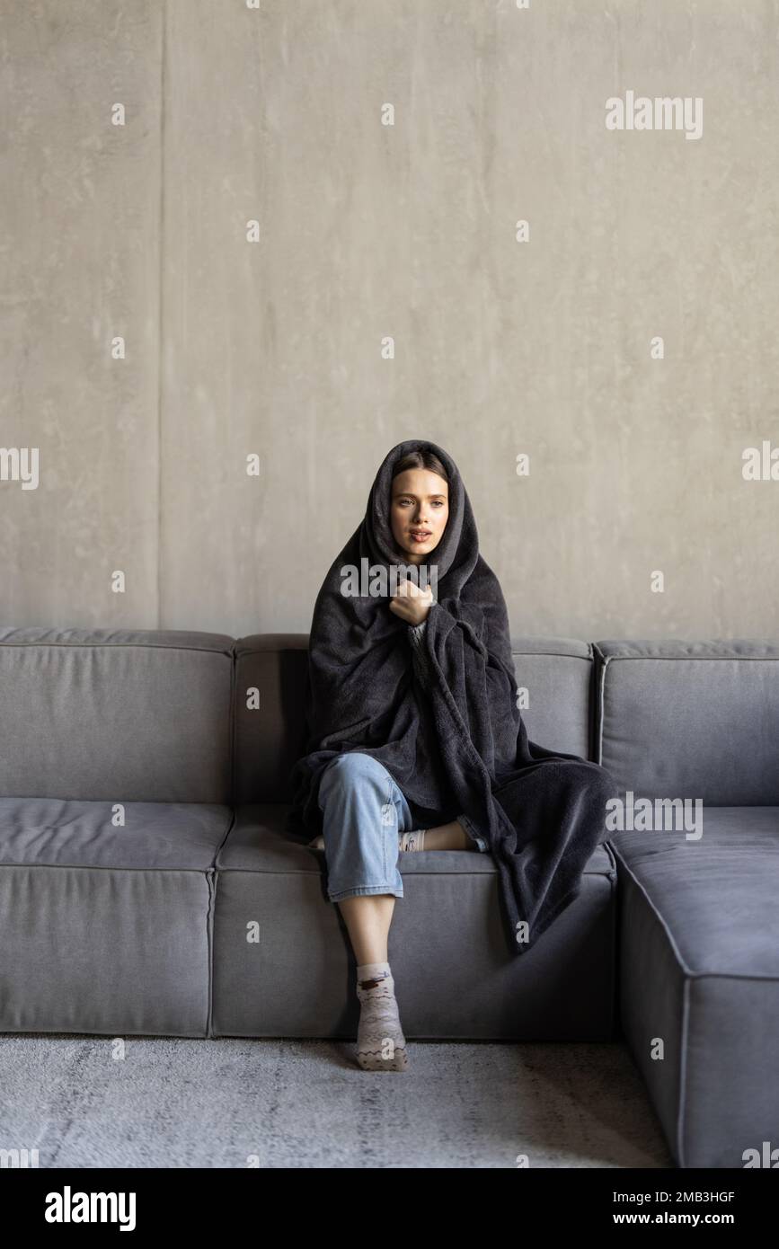 woman snuggling into a warm blanket while sitting on her sofa with a ...