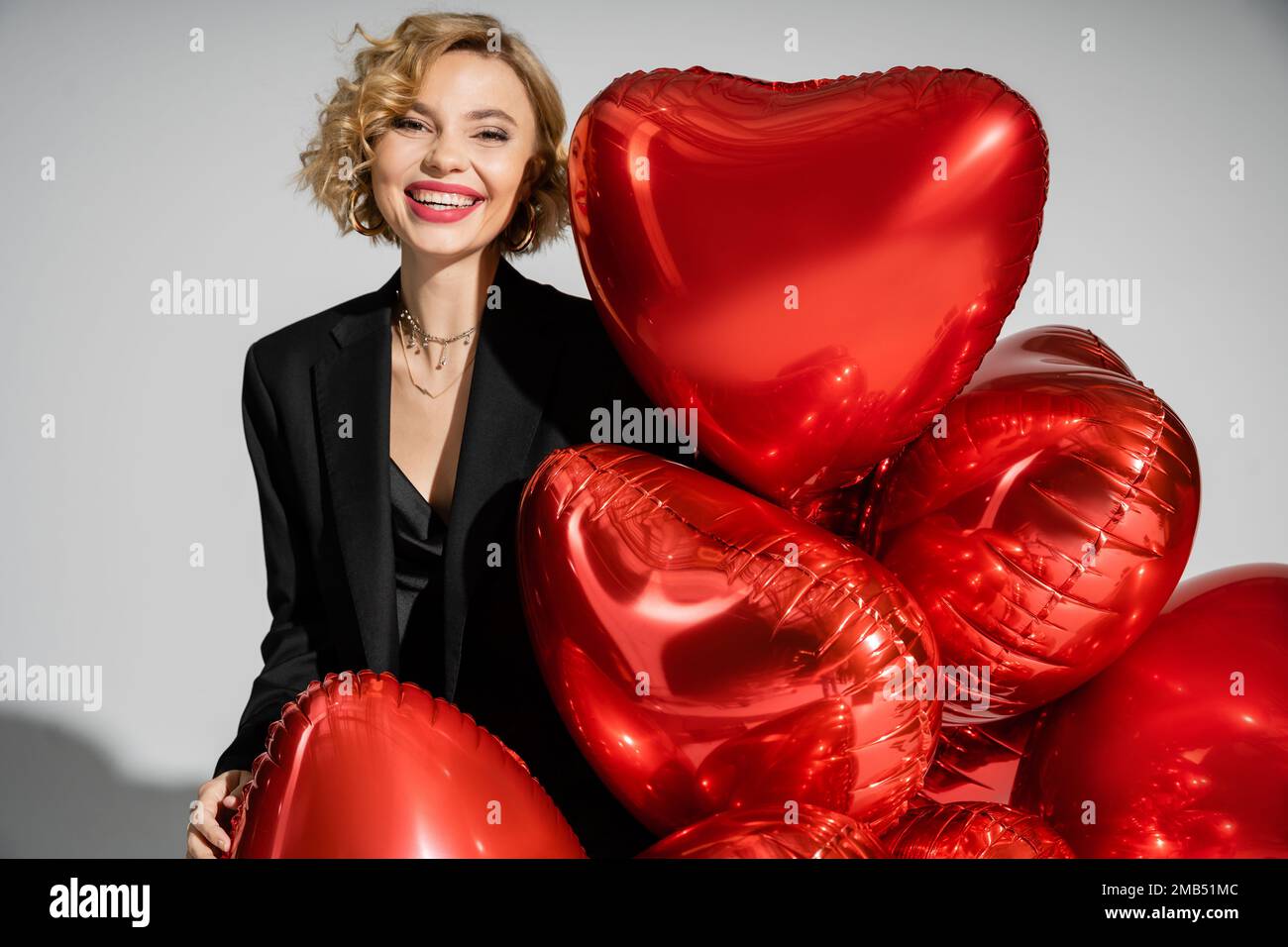 happy young woman in black slip dress and blazer smiling near red heart ...
