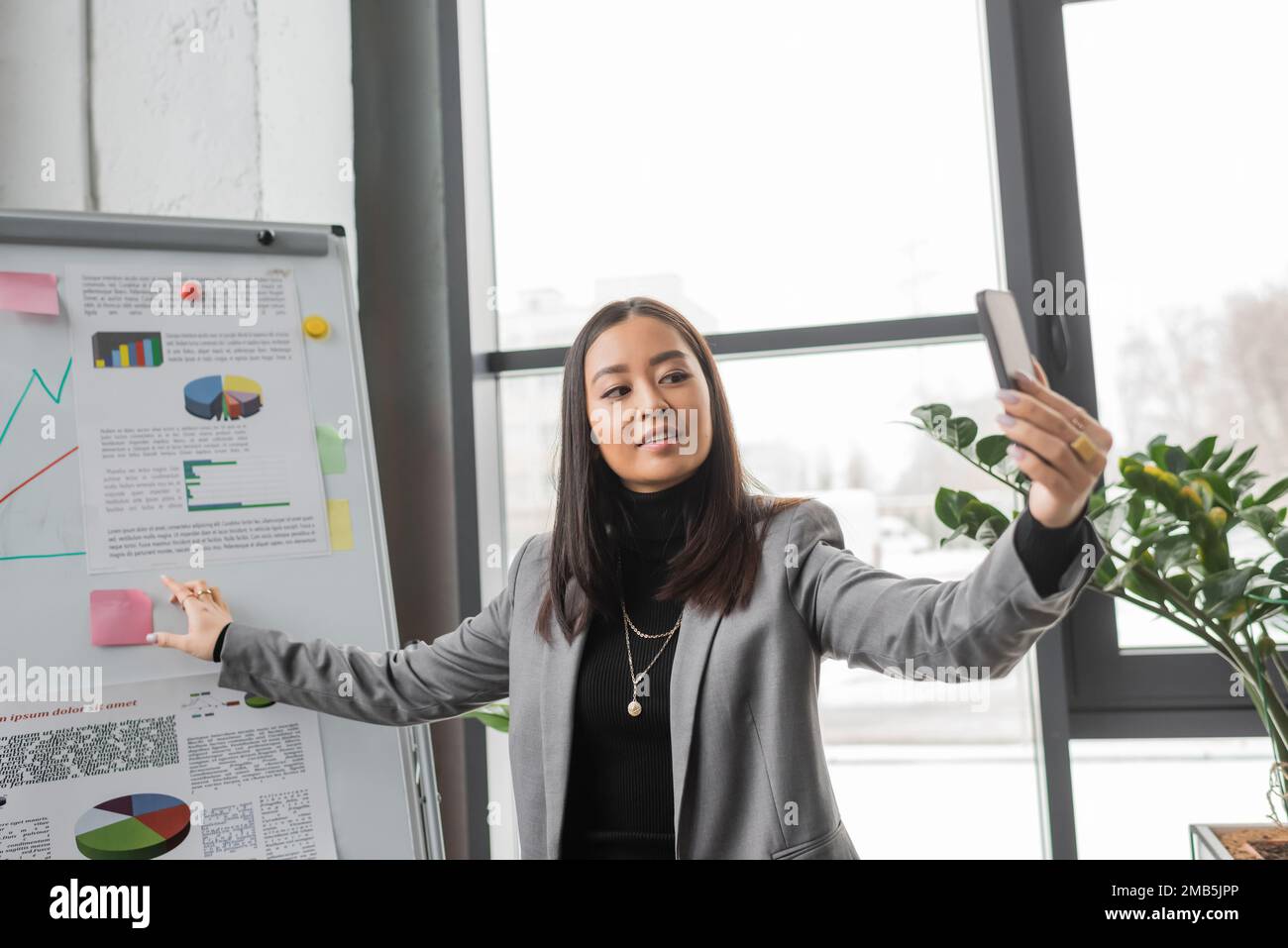 Asian designer pointing at flip chart during video call on smartphone ...