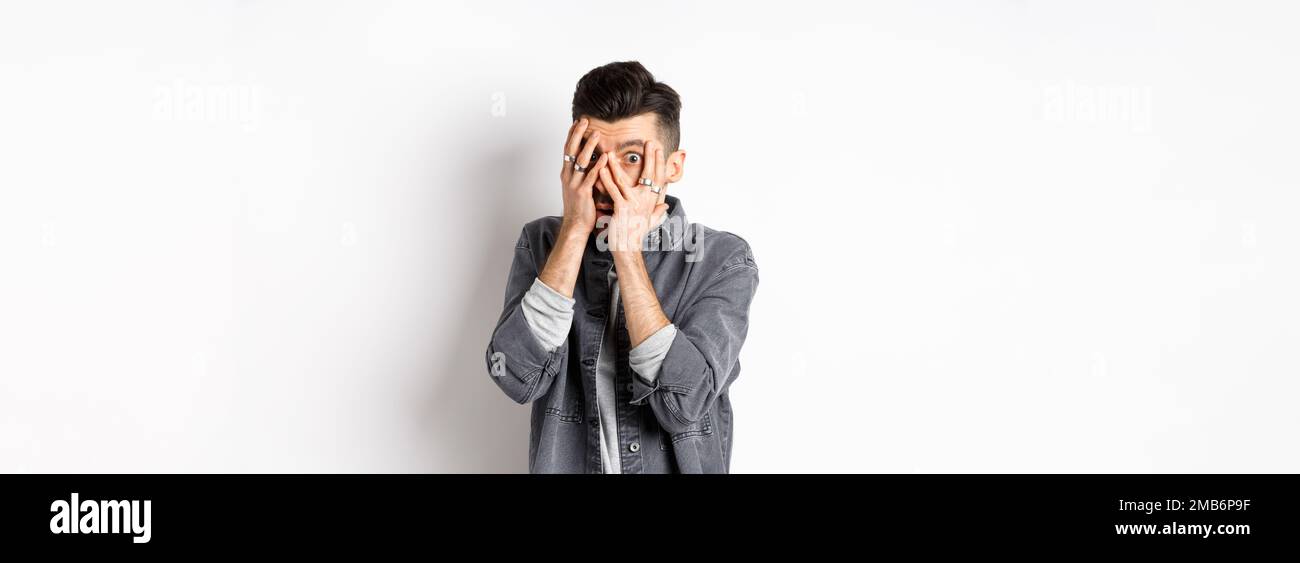 Scared young man peeking through fingers with nervous face, staring at ...