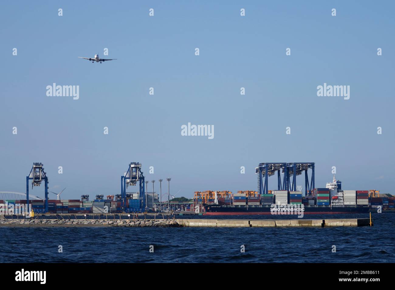 An airplane flying over cranes, containers and a shipping vessel ...