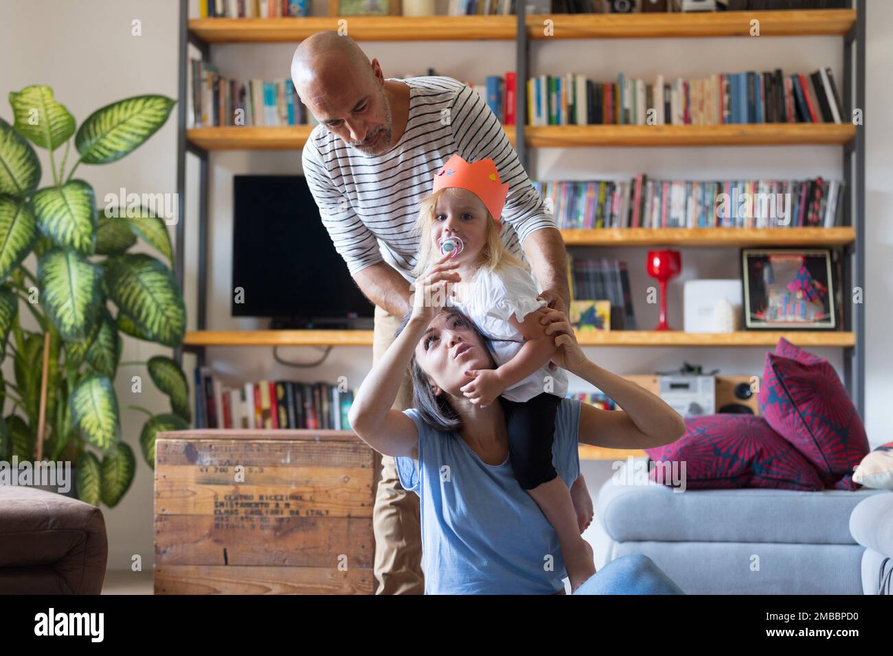 little baby girl with wreath on her head rides piggyback on mom's shoulders holding her pacifier ...