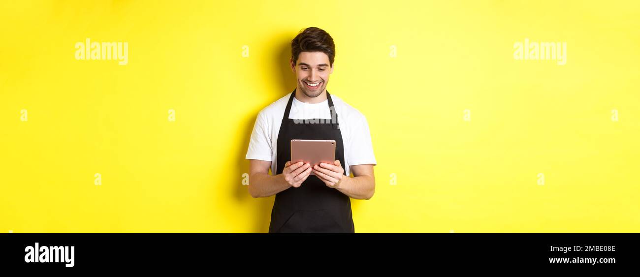 Seller in black apron looking at digital tablet screen, smiling pleased ...