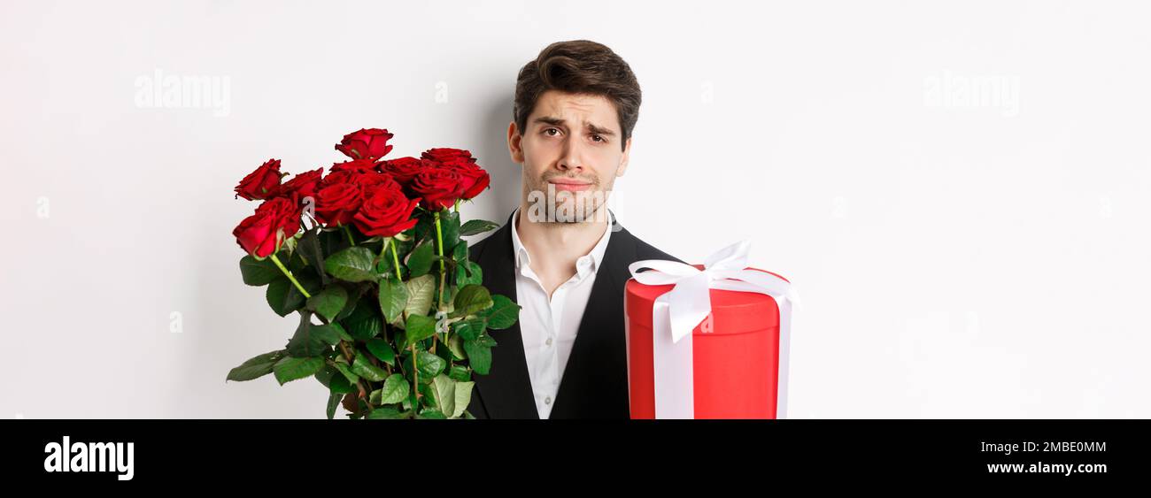 Close-up of sad man in suit, holding bouquet of red roses and a gift ...