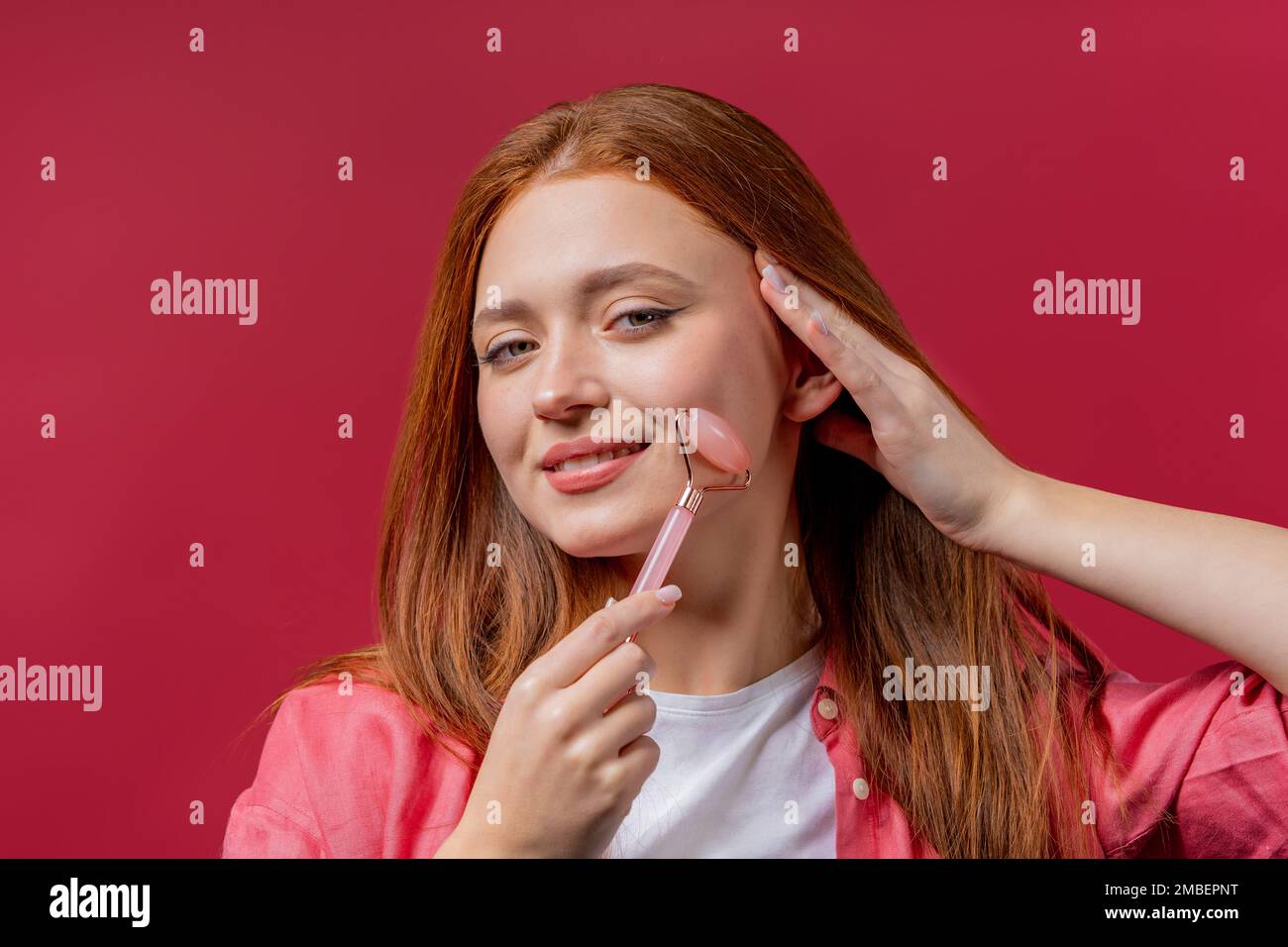 Ginger woman doing face massage with rose quartz stone roller on pink ...