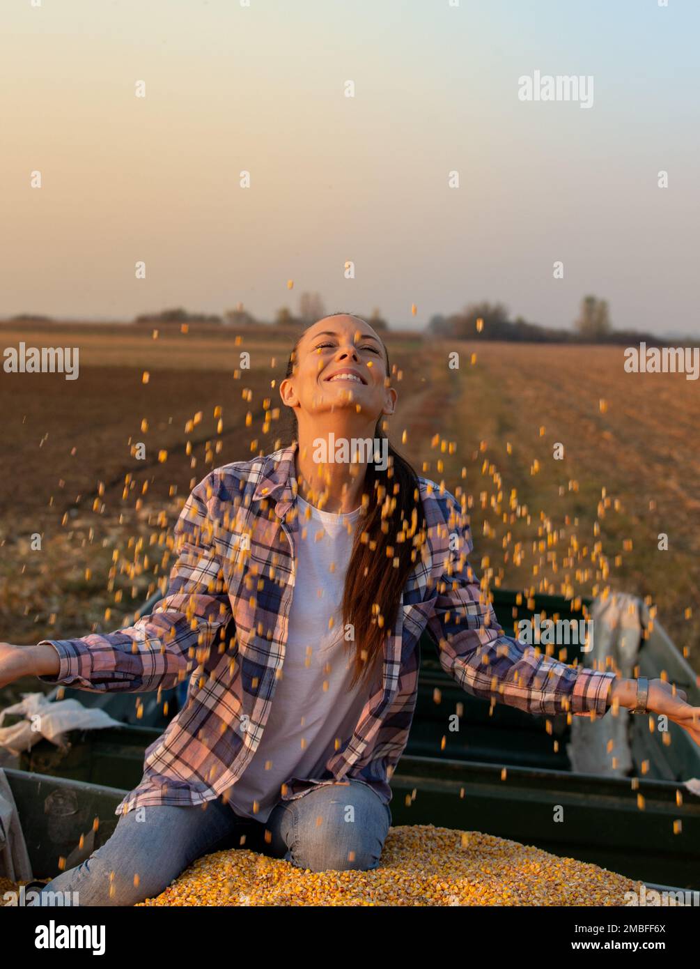 Happy farmer woman sitting on corn pile in tractor trailer nd throwing ...